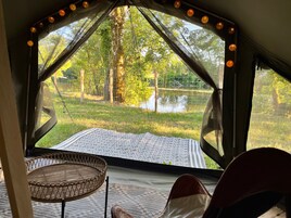 Tenda com vista para uma área gramada e um lago. No interior, há uma cadeira de vime e um tapete estampado.