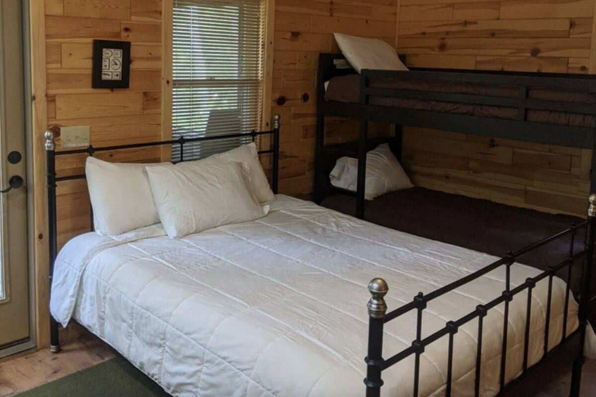 Uma cama de solteiro com colcha branca num quarto com paredes de madeira e uma cama beliche em cima.