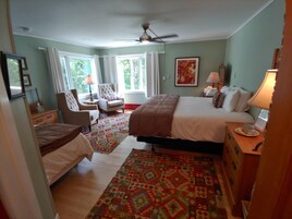 Quarto com cama, duas cadeiras, escrivaninha, luminária e janela com cortinas.