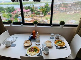 Mesa de café da manhã com pratos de comida, uma garrafa e xícaras, em frente a uma janela com vista para a cidade.