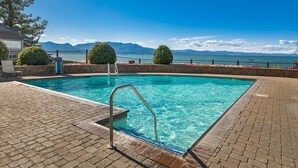 Uma piscina retangular com água azul cristalina, ladeada por um deck de tijolos e um guarda-corpo de metal. Ao fundo, vê-se uma vista de montanhas e um corpo de água.