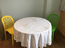 Mesa redonda com toalha branca e detalhes em renda, rodeada por duas cadeiras, uma amarela e outra verde.