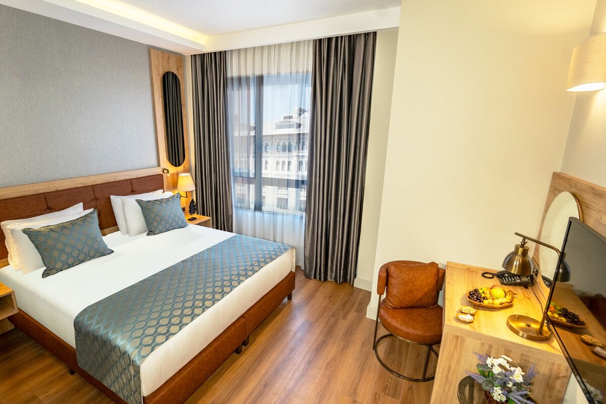 Quarto de hotel com uma cama grande, uma escrivaninha com computador e abajur, uma cadeira e vista para edifícios pela janela.