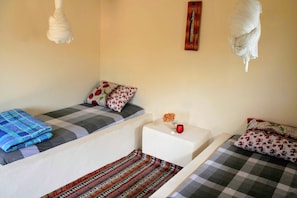 Quarto pequeno e simples com duas camas de solteiro, uma mesinha de cabeceira branca e uma pequena mesa com uma vela vermelha.