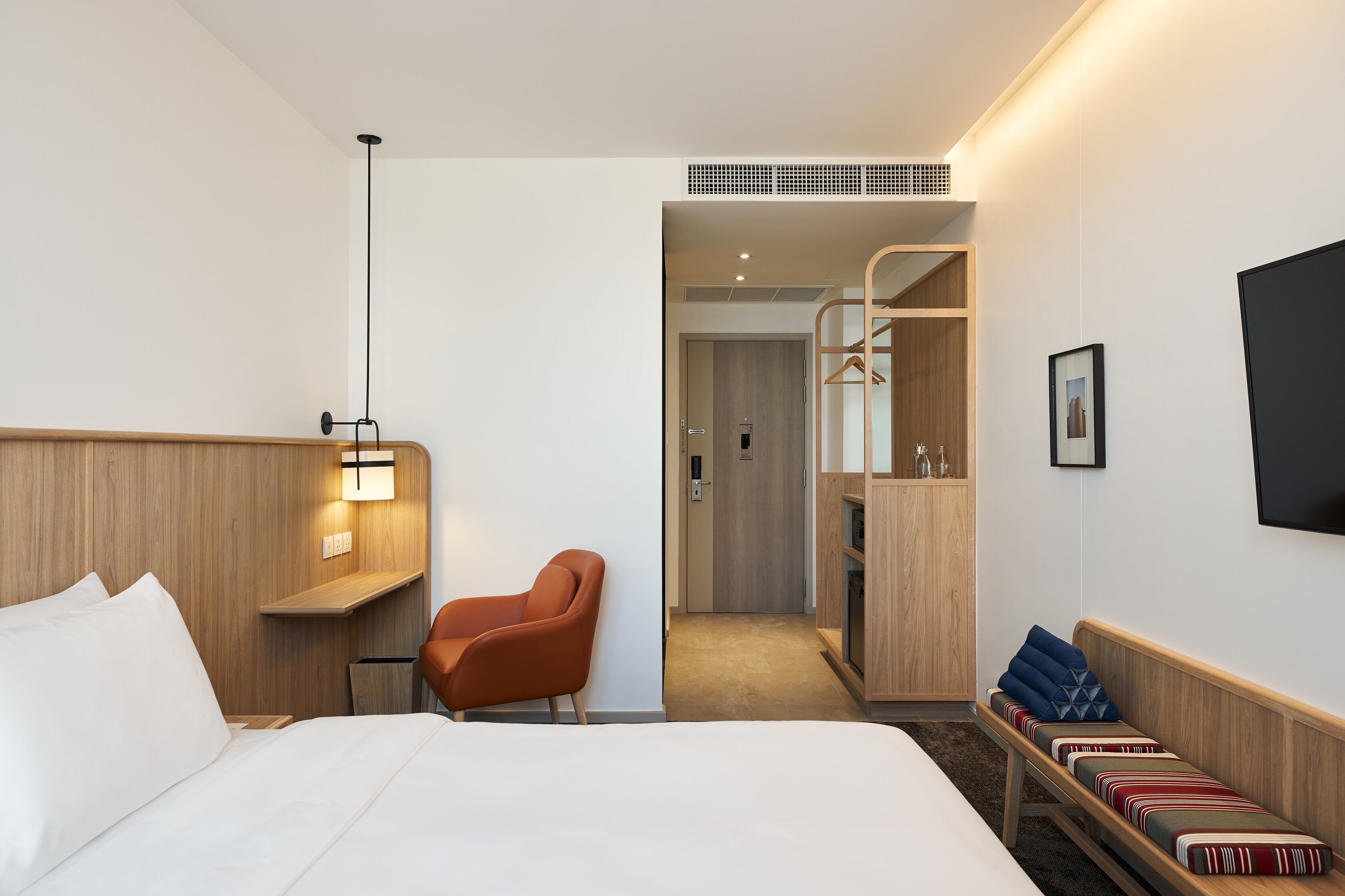 Quarto de hotel com uma cama, uma cadeira, um banco, um guarda-roupa e uma televisão.