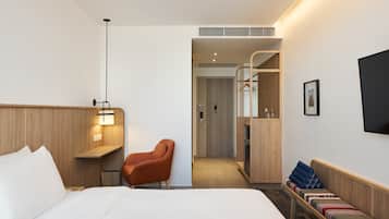 Quarto de hotel com uma cama, uma cadeira, um banco, um guarda-roupa e uma televisão.