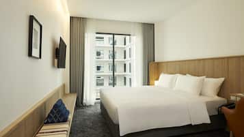 Quarto de hotel com uma cama grande, um banco com almofadas, televisão e janela com cortinas.