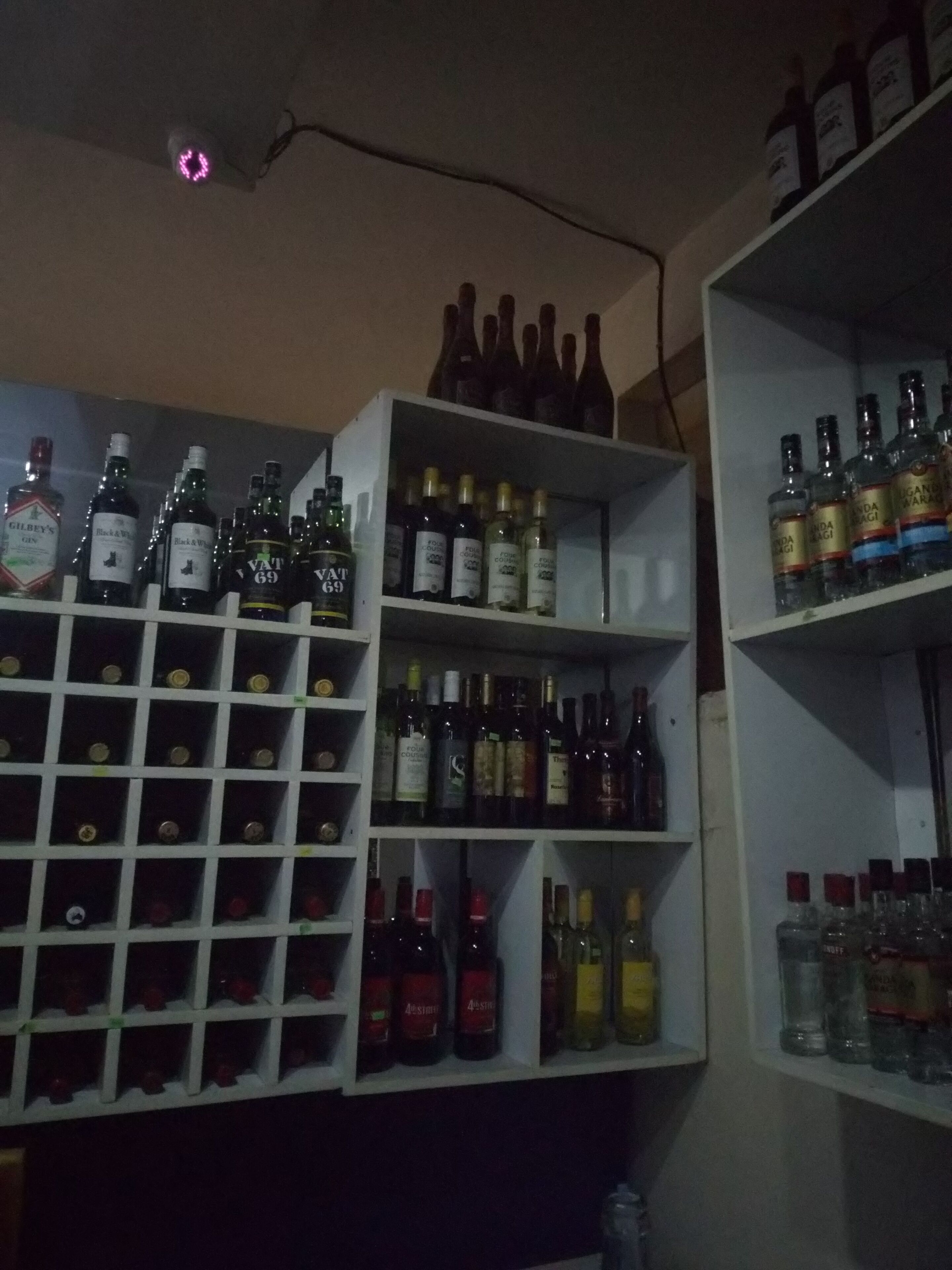 Prateleiras repletas de diversas garrafas de bebidas alcoólicas, incluindo marcas como Gilbey's Gin e Four Corners.