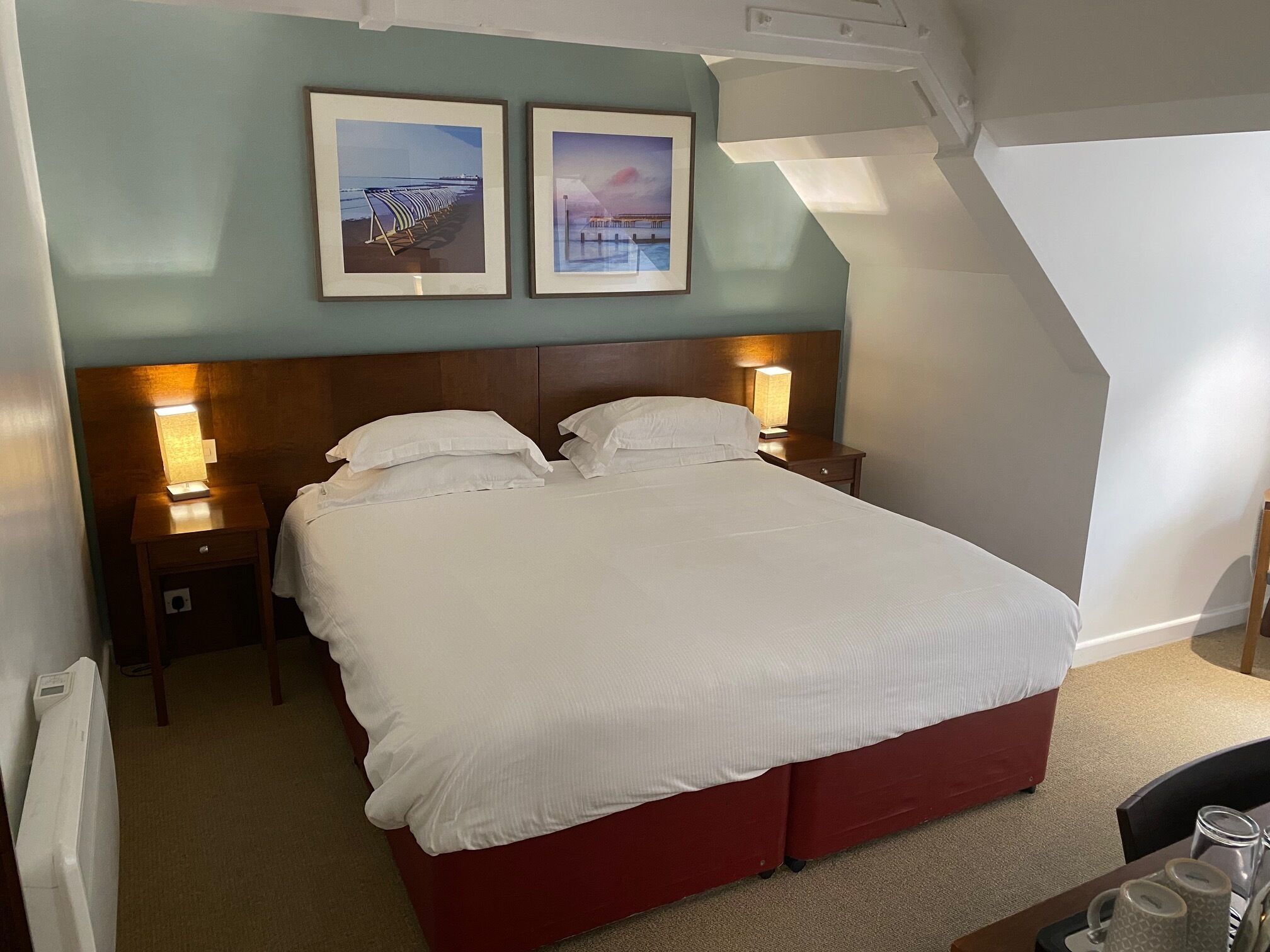 Quarto de hotel com uma cama grande, duas mesas de cabeceira com luminárias, uma cabeceira e duas quadros na parede.