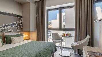 Quarto de hotel com cama, uma pequena mesa com cadeira, vista da cidade através de grandes janelas e um mural na parede.