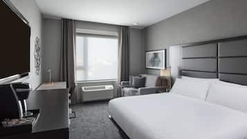 Quarto de hotel moderno com uma cama grande, uma escrivaninha com cafeteira, uma cadeira, uma televisão e uma janela ampla com cortinas.