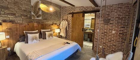 Quarto com cama grande, cabeceira de madeira, parede de pedra, luminária pendente e uma cadeira de vime.