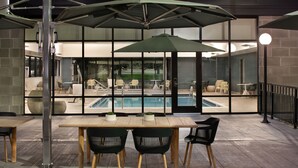 Uma área externa com mesas e cadeiras, vista para a piscina e um guarda-sol coberto.
