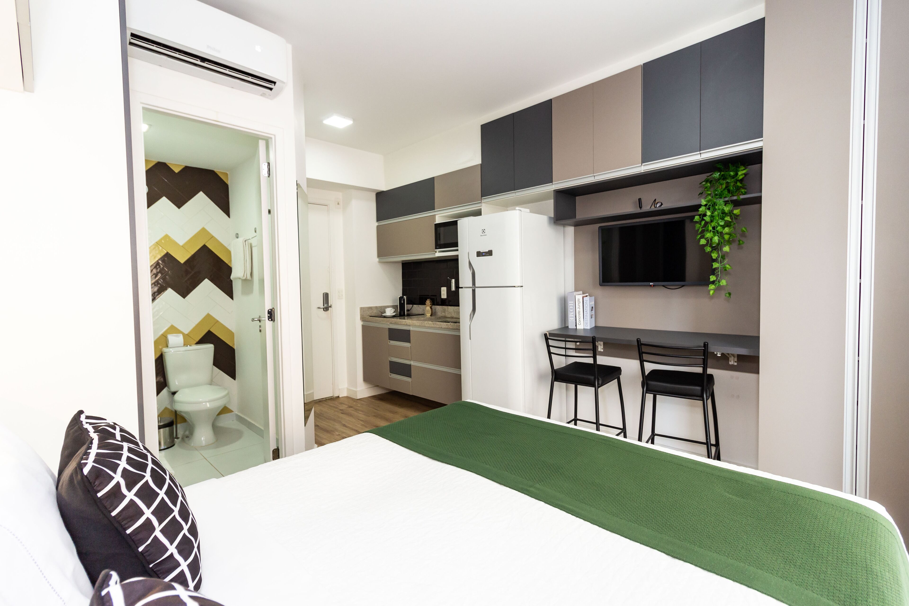 Quarto de hotel compacto com cama, pequena área de cozinha, banheiro e sala de jantar.