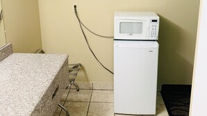 Quarto pequeno e compacto com cozinha compacta, incluindo micro-ondas e frigorífico. Há uma bancada com pia e suporte para toalhas. O quarto tem chão em azulejos e uma porta que dá para outra área.