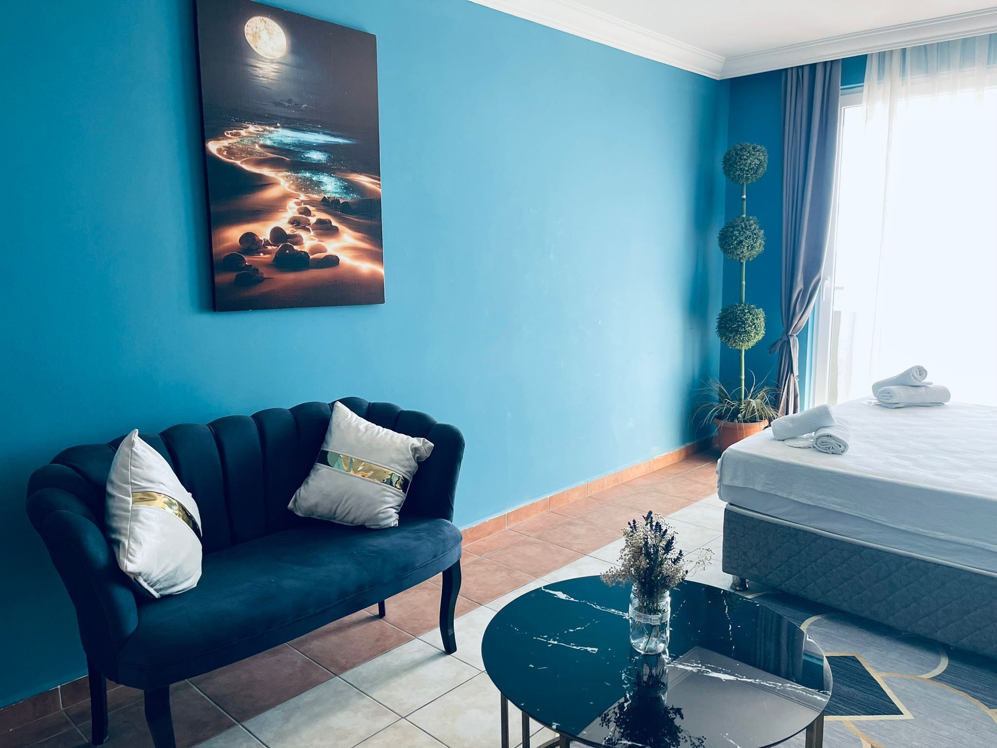 Quarto com parede azul, cama com roupa de cama branca, sofá azul escuro com almofadas brancas, mesa pequena com um vaso de flores e uma pintura de uma cena de praia.