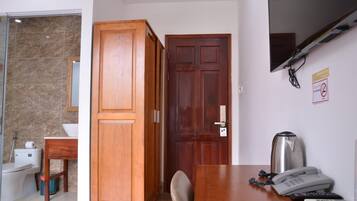 Quarto de hotel com guarda-roupa de madeira, mesa com telefone, chaleira e televisão fixada na parede.