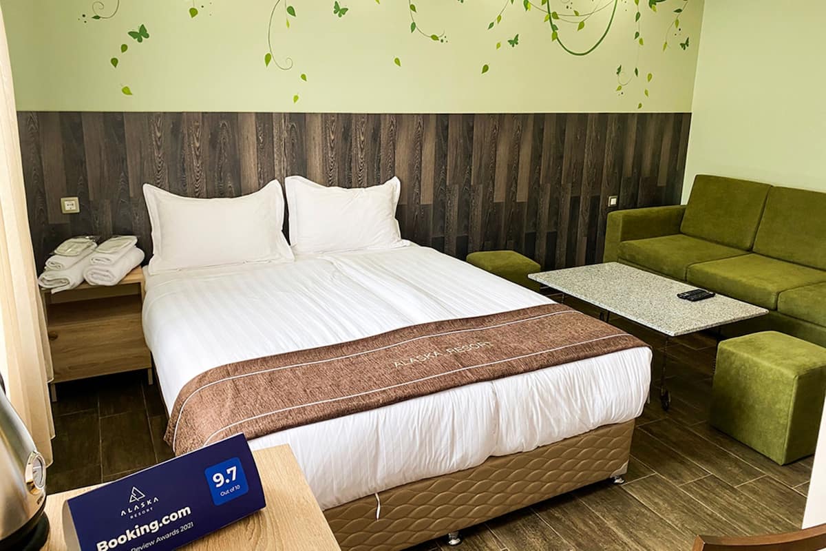 Quarto de hotel com uma cama, um sofá verde, uma pequena mesa e uma parede com um desenho de folha. Um sinal na mesa indica uma avaliação de 9,7 em 10.