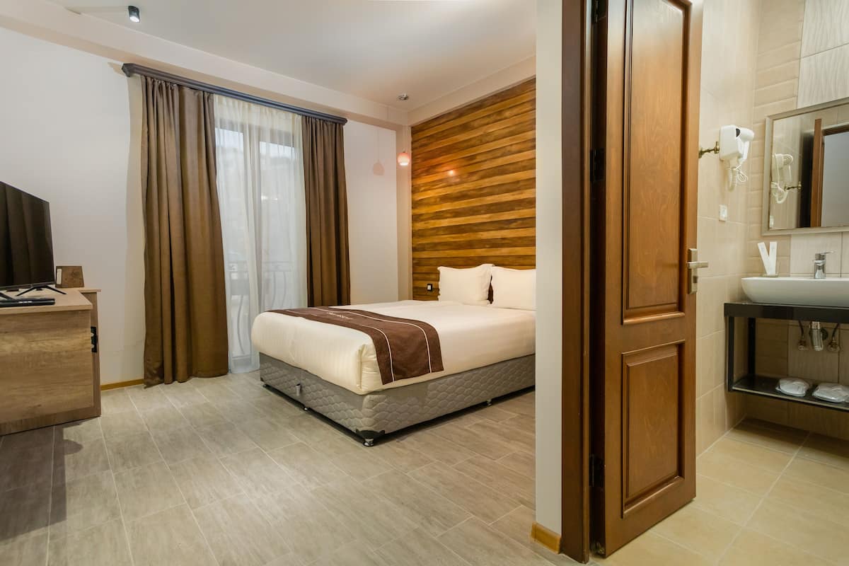 Quarto de hotel com cama, cabeceira de madeira, televisão, escrivaninha e banheiro com pia e espelho.