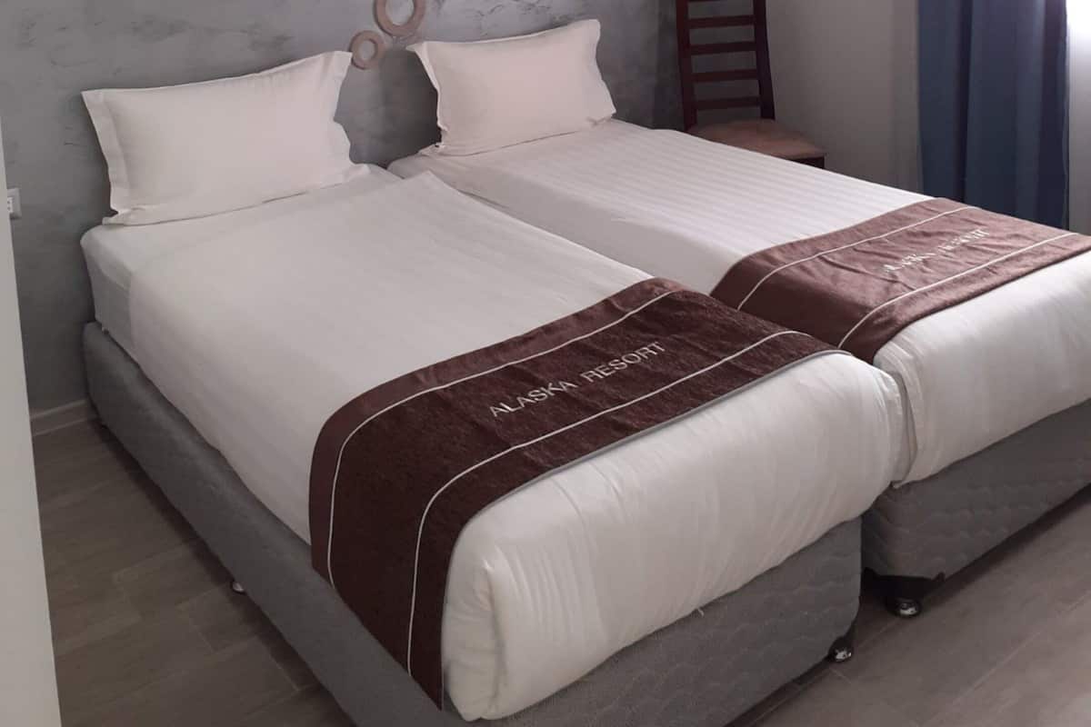 Cama de casal com cabeceira cinza, roupa de cama branca e manta marrom decorativa. Há um enfeite circular na parede e uma luminária pendente.
