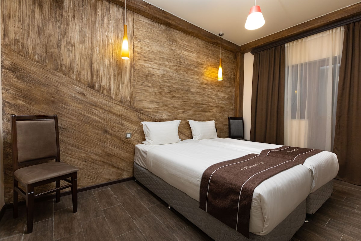 Quarto de hotel com uma cama grande, painel de parede em madeira, uma cadeira e uma janela com cortinas.