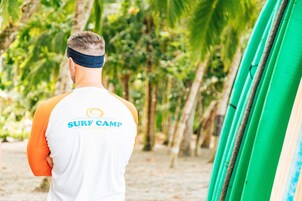 Uma pessoa vestindo camisa branca com a inscrição 'SURF CAMP' e 'COSTA RICA' nas costas, em um ambiente natural com árvores e um caminho de areia.
