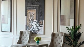 Um hall com duas poltronas estofadas, uma mesa pequena com um vaso, e uma grande obra de arte abstrata na parede.