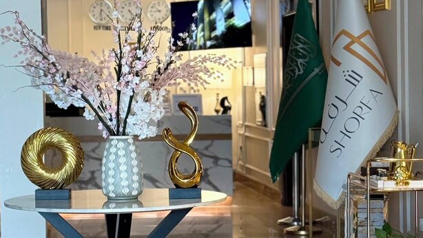 Uma área de recepção com mesa de vidro, um vaso com flores, um objeto decorativo dourado e uma bandeira.