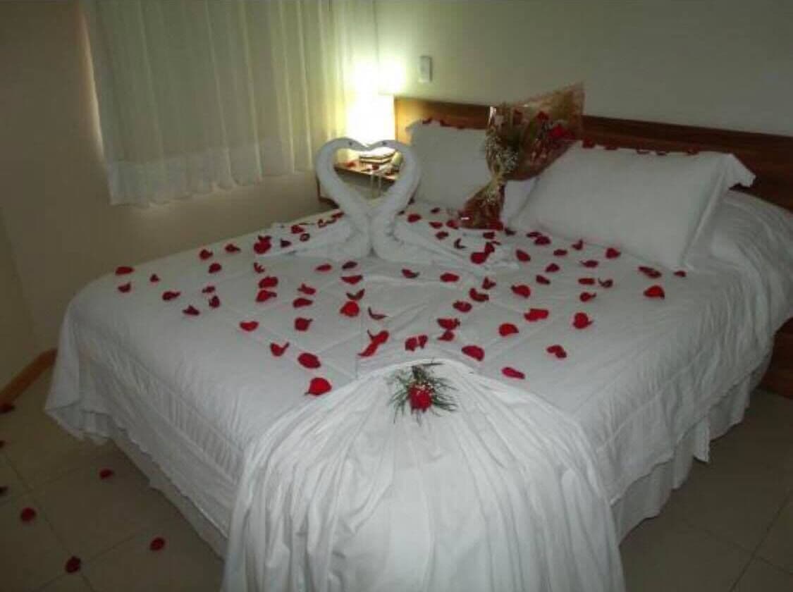 Uma cama com pétalas de rosa e uma decoração em forma de coração, um cobreleiro branco e uma mesa de cabeceira com abajur.