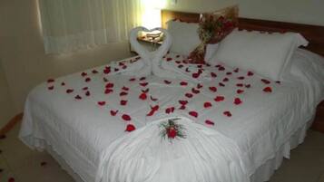 Uma cama com pétalas de rosa e uma decoração em forma de coração, um cobreleiro branco e uma mesa de cabeceira com abajur.