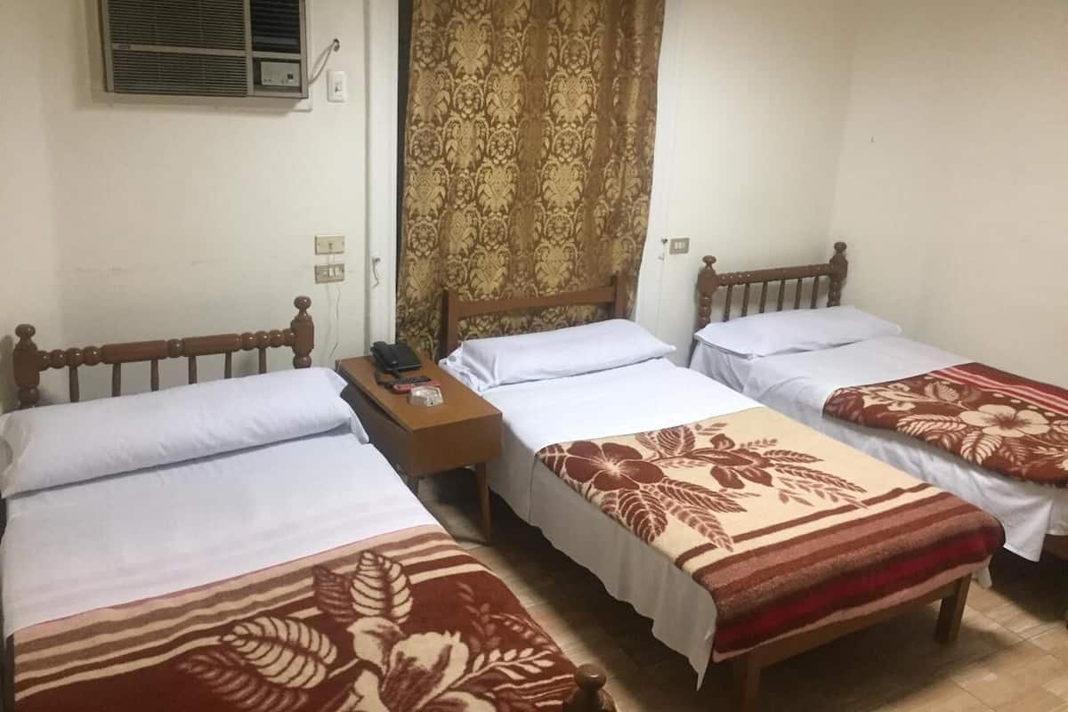 Quarto com três camas de solteiro, cada uma com cabeceira de madeira e colcha estampada. Um criado-mudo de madeira com telefone e luminária está presente em um dos lados. Um ar-condicionado fixado na parede é visível acima da cama à esquerda.