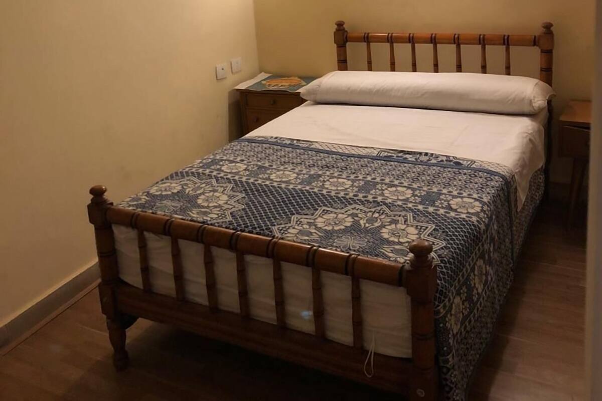 Quarto com cama de madeira, mesa de cabeceira, luminária na parede e quadro emoldurado.