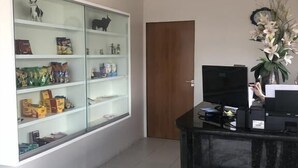 Uma área de recepção com balcão de mármore preto, uma vitrine de vidro com lanches, uma porta de madeira e uma TV fixada na parede.