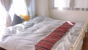 Uma cama com colcha estampada e um corredor decorativo, uma janela com cortinas translúcidas e uma parede com quadros emoldurados.