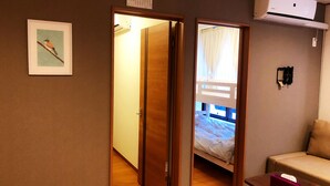 Quarto com porta de madeira que dá para um quarto com beliche, uma mesinha com caixa de lenços e um ar condicionado na parede.