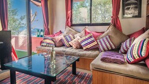 Uma sala de estar aconchegante com sofá, almofadas coloridas, uma mesa de centro de vidro e vista para o verde lá fora.