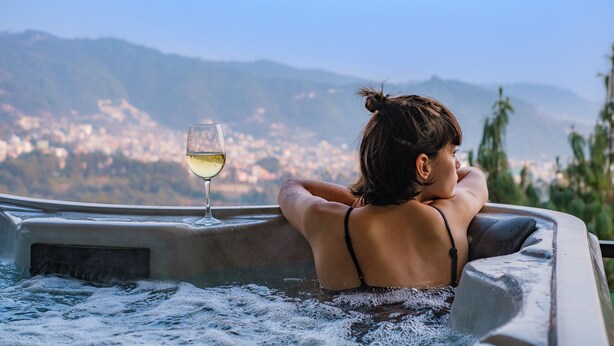 Uma pessoa relaxando em uma banheira de hidromassagem, apreciando uma vista panorâmica da cidade e montanhas, com uma taça de vinho.