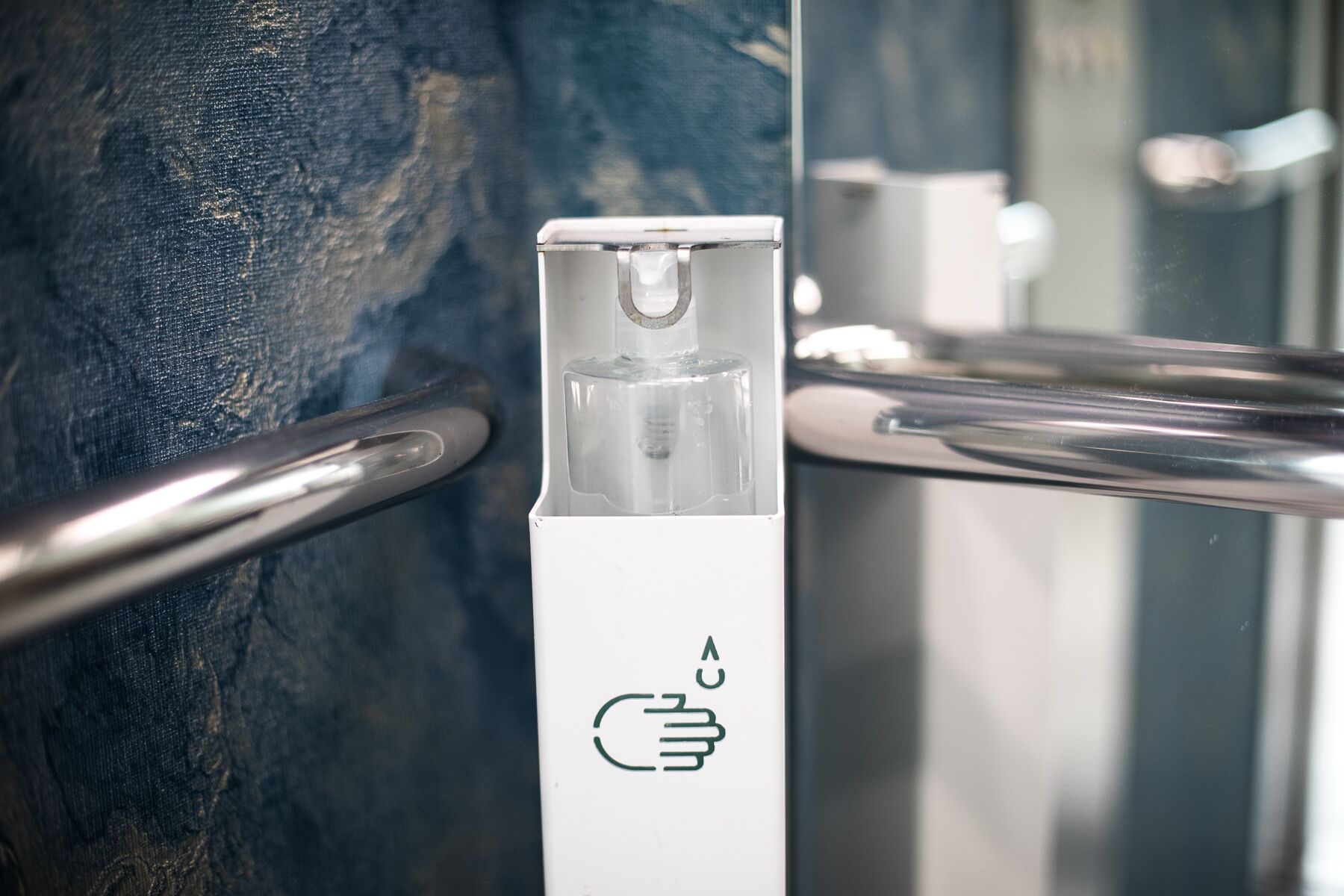 Um dispensador de álcool em gel fixado em um corrimão de metal, com uma parede texturizada ao fundo.