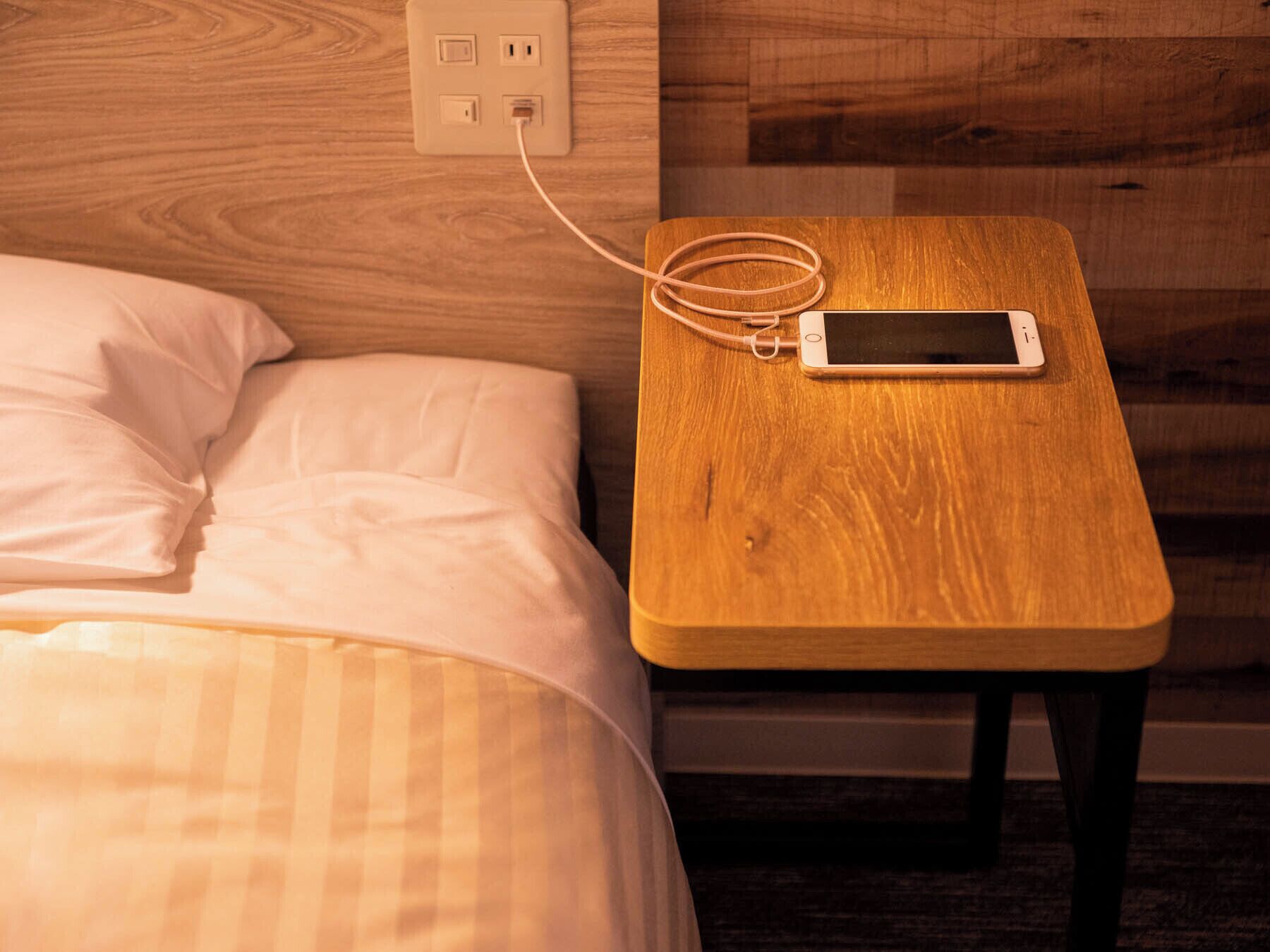 Cama com lençóis brancos, mesa de cabeceira de madeira com um smartphone e cabo de carregamento, e uma tomada na parede.