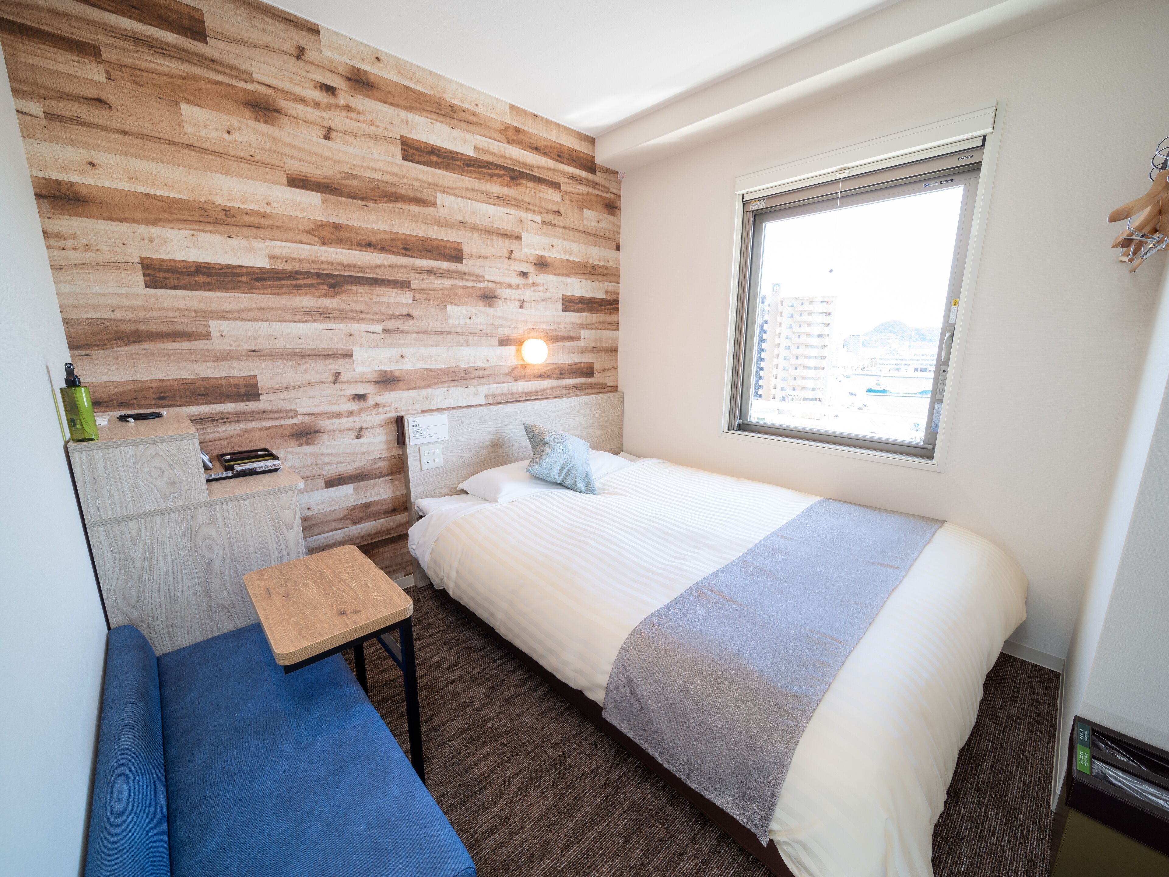 Quarto de hotel compacto com parede de madeira, uma cama de solteiro, uma pequena escrivaninha, um banco azul e uma janela com vista para a cidade.