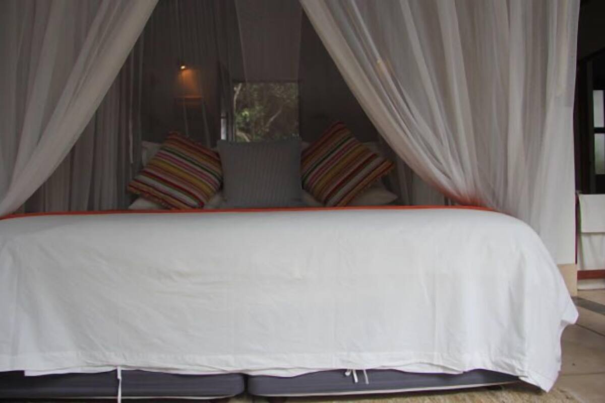Cama com roupa de cama branca e almofadas listradas, coberta por cortinas finas.