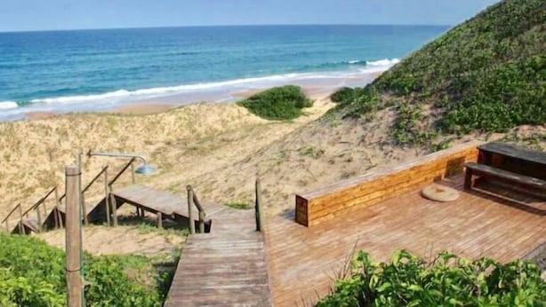 Um deck de madeira com um banco e um flutuador, com vista para uma praia com dunas de areia e o oceano.