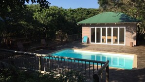 Uma área de piscina com deck de madeira, um pequeno edifício com telhado verde e uma piscina cercada.