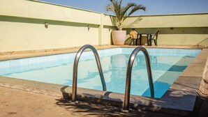 Piscina retangular com grades de metal, uma palmeira e uma mesa com cadeiras ao fundo.
