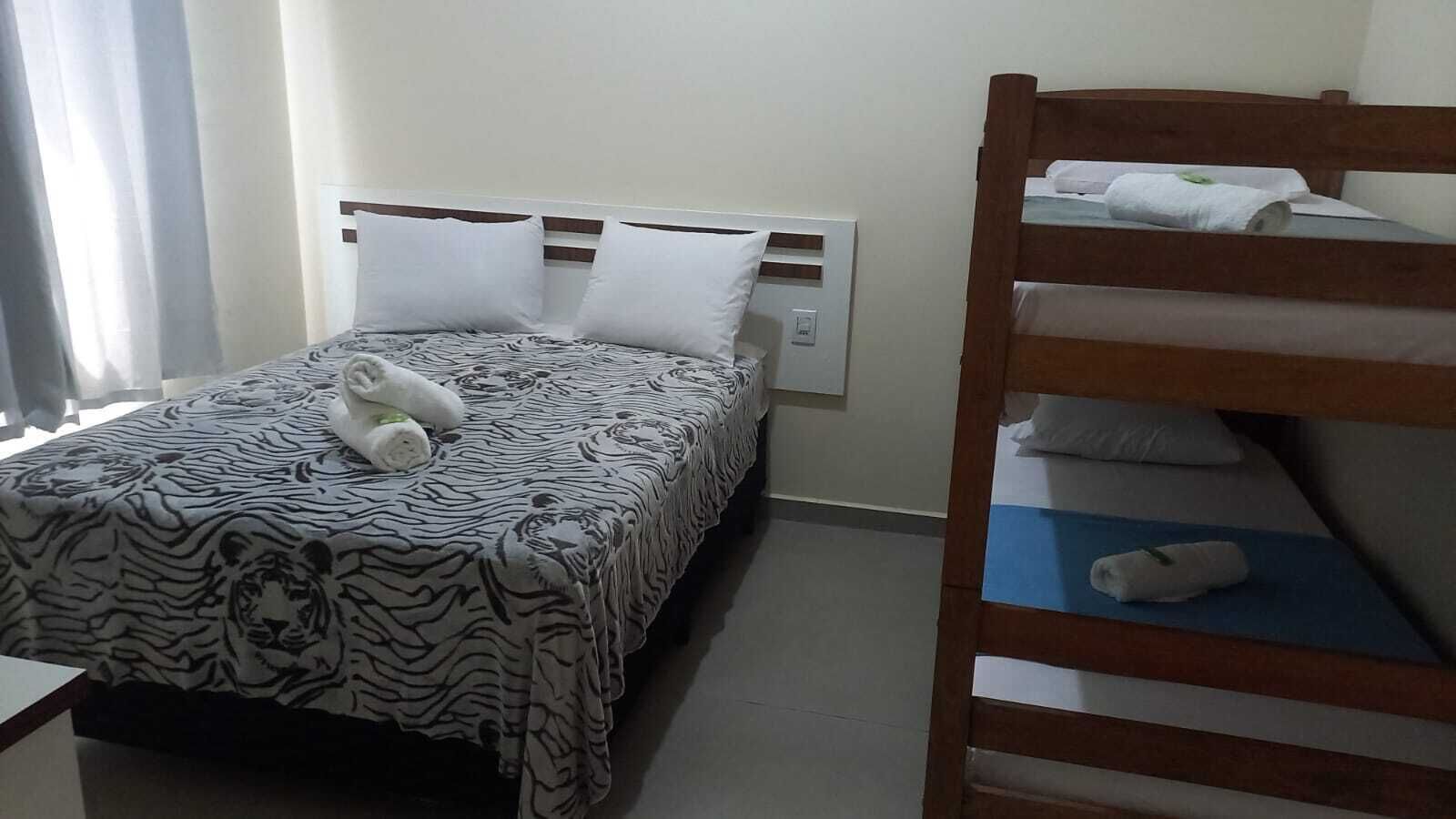 Uma cama de solteiro com colcha estampada em padrão de zebra e duas almofadas, uma mesinha de cabeceira e uma cama beliche de madeira à direita.