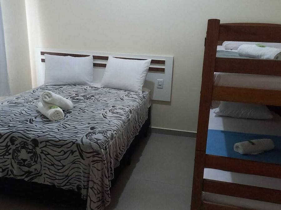 Uma cama de solteiro com colcha estampada em padrão de zebra e duas almofadas, uma mesinha de cabeceira e uma cama beliche de madeira à direita.