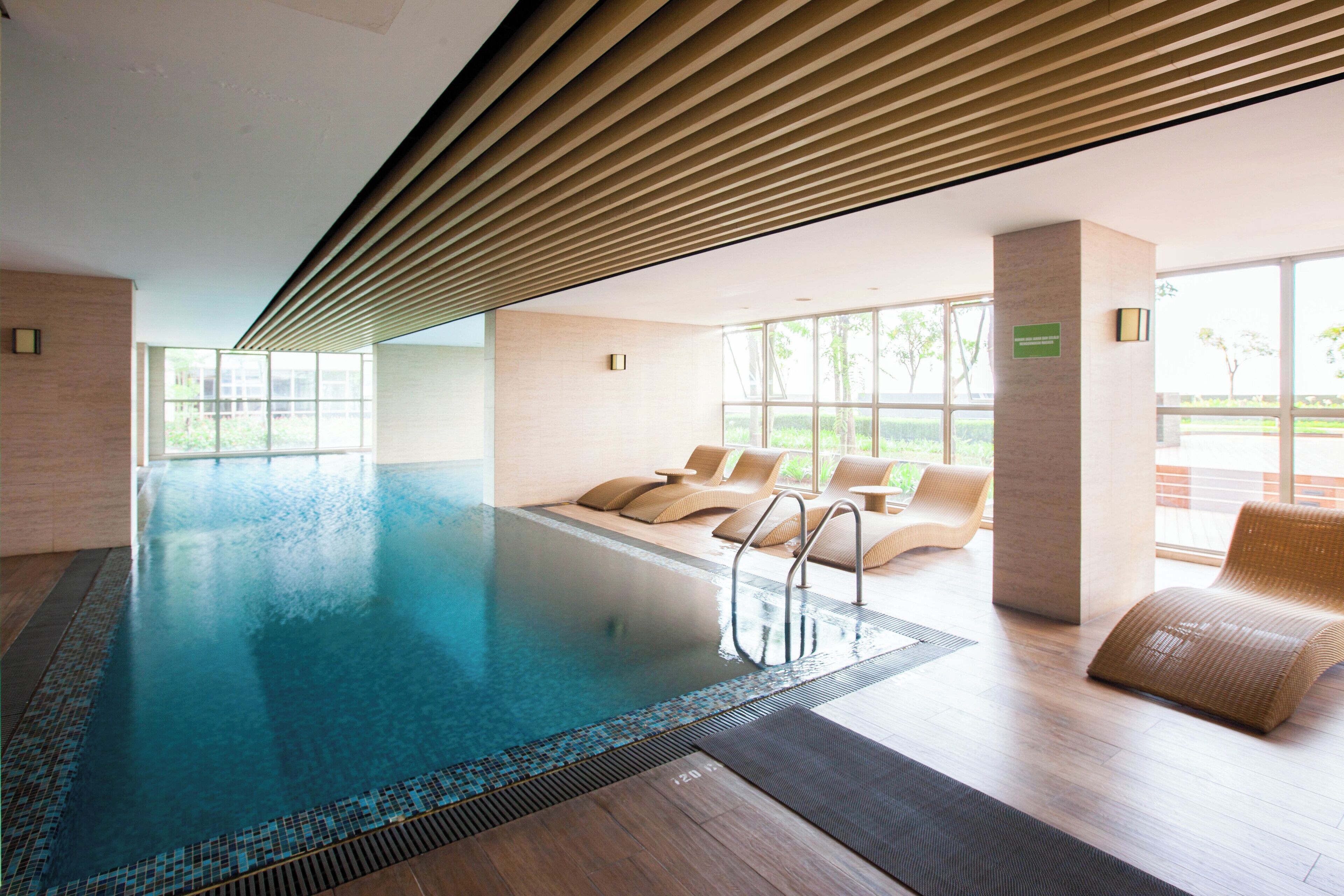 Piscina interior moderna com paredes de vidro transparente, teto de madeira e confortáveis espreguiçadeiras.