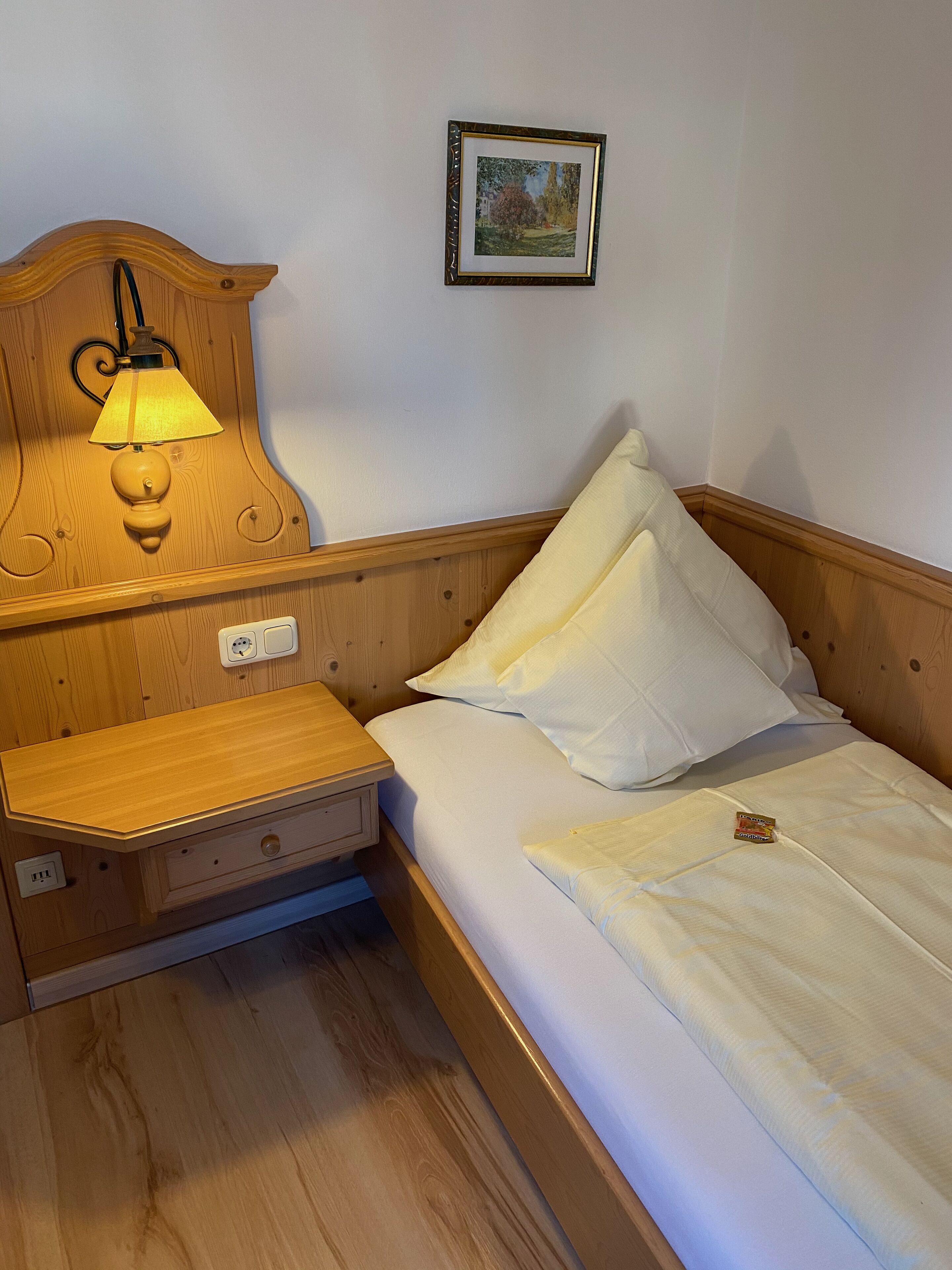Uma cama de madeira com roupa de cama branca, uma mesinha de cabeceira, um abajur e um quadro na parede.