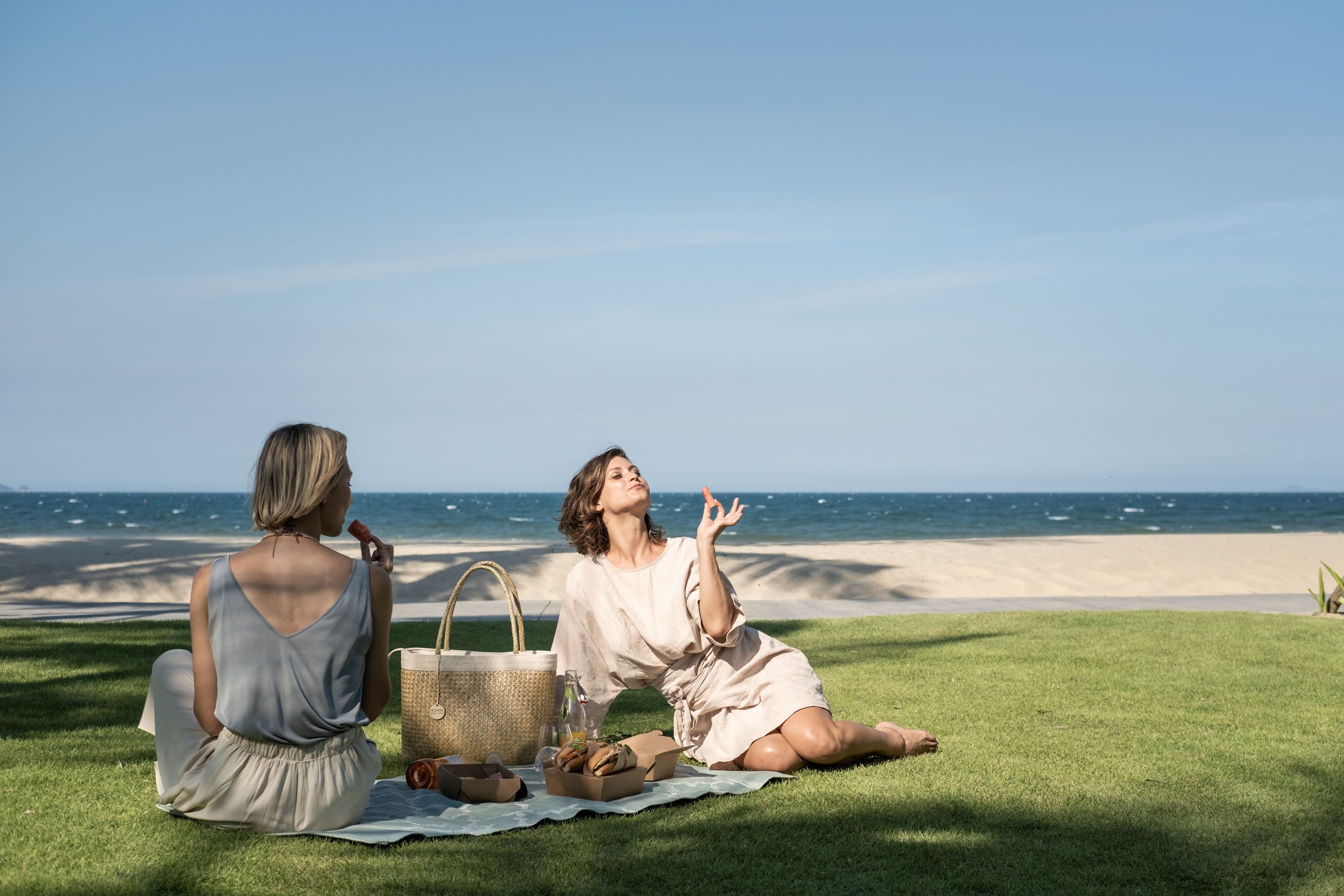Duas pessoas sentadas em uma toalha na praia, com uma bolsa de praia e uma garrafa de água no chão.