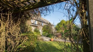 Uma casa rústica com telhado de palha, rodeada por um jardim exuberante e uma pérgola de madeira.